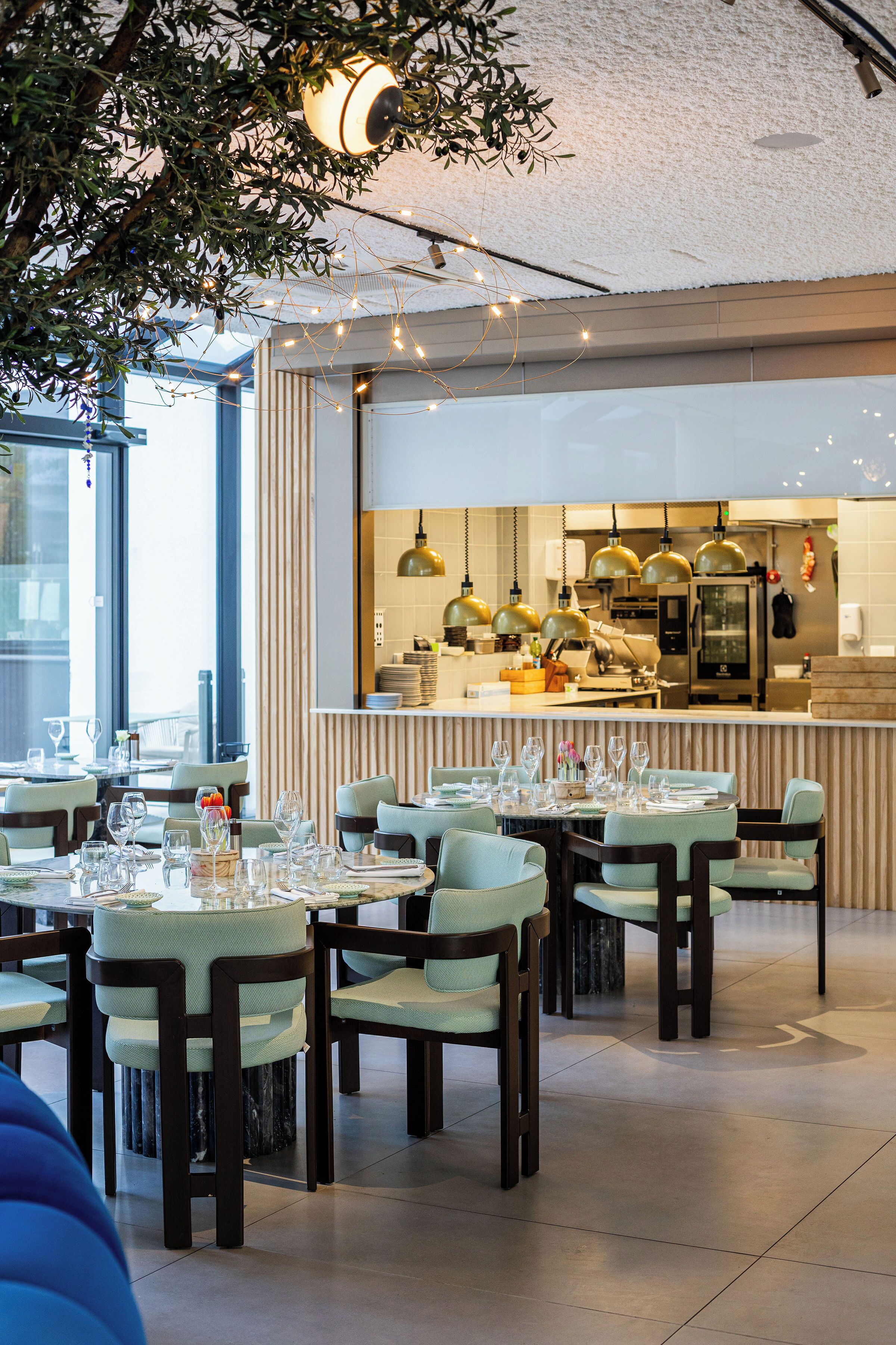 Restaurante com mesas preparadas para refeições, cozinha ao fundo e uma grande janela que permite entrada de luz natural.