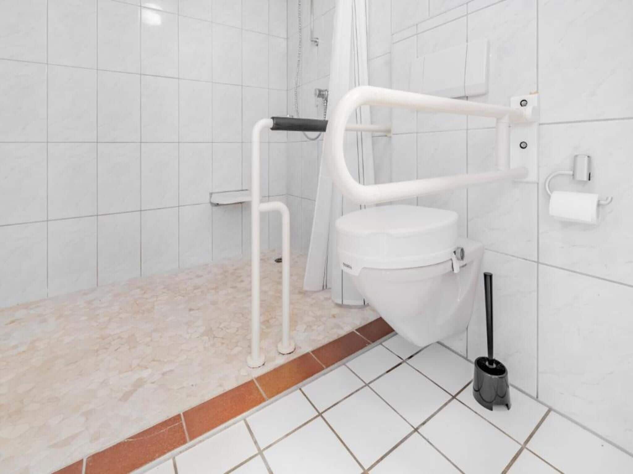 Casa de banho com sanita branca, zona de duche e barra de apoio.