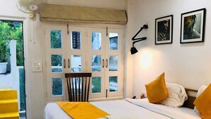 Quarto com cama, cadeira, escrivaninha e janela com vista para vegetação.