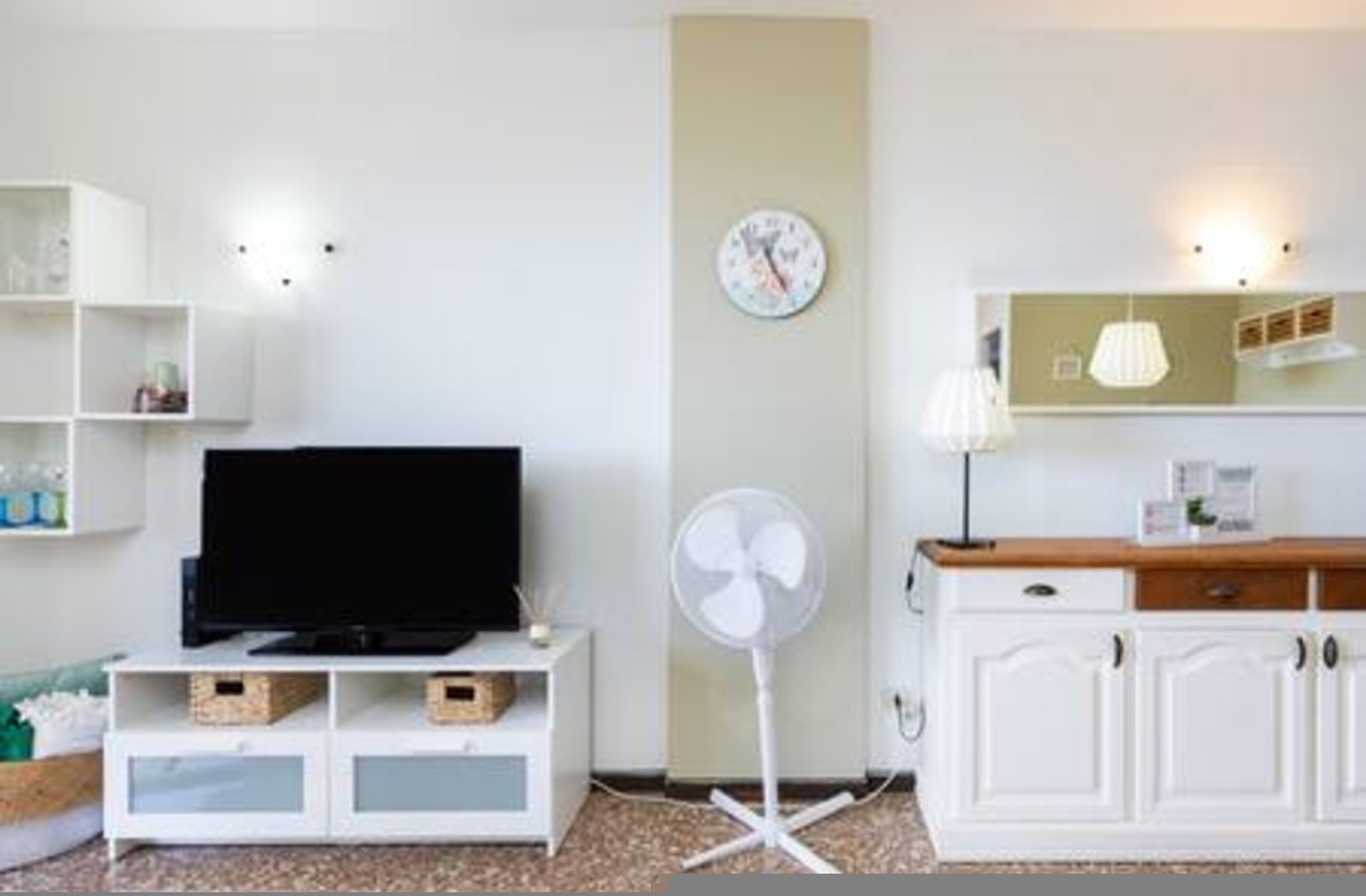 Uma sala de estar moderna com uma televisão de tela plana sobre um armário branco, um ventilador de chão e um armário de madeira com gavetas.