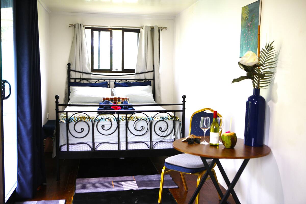 Um quarto com cama, uma mesinha com cadeira, uma janela com cortinas e um quadro na parede.