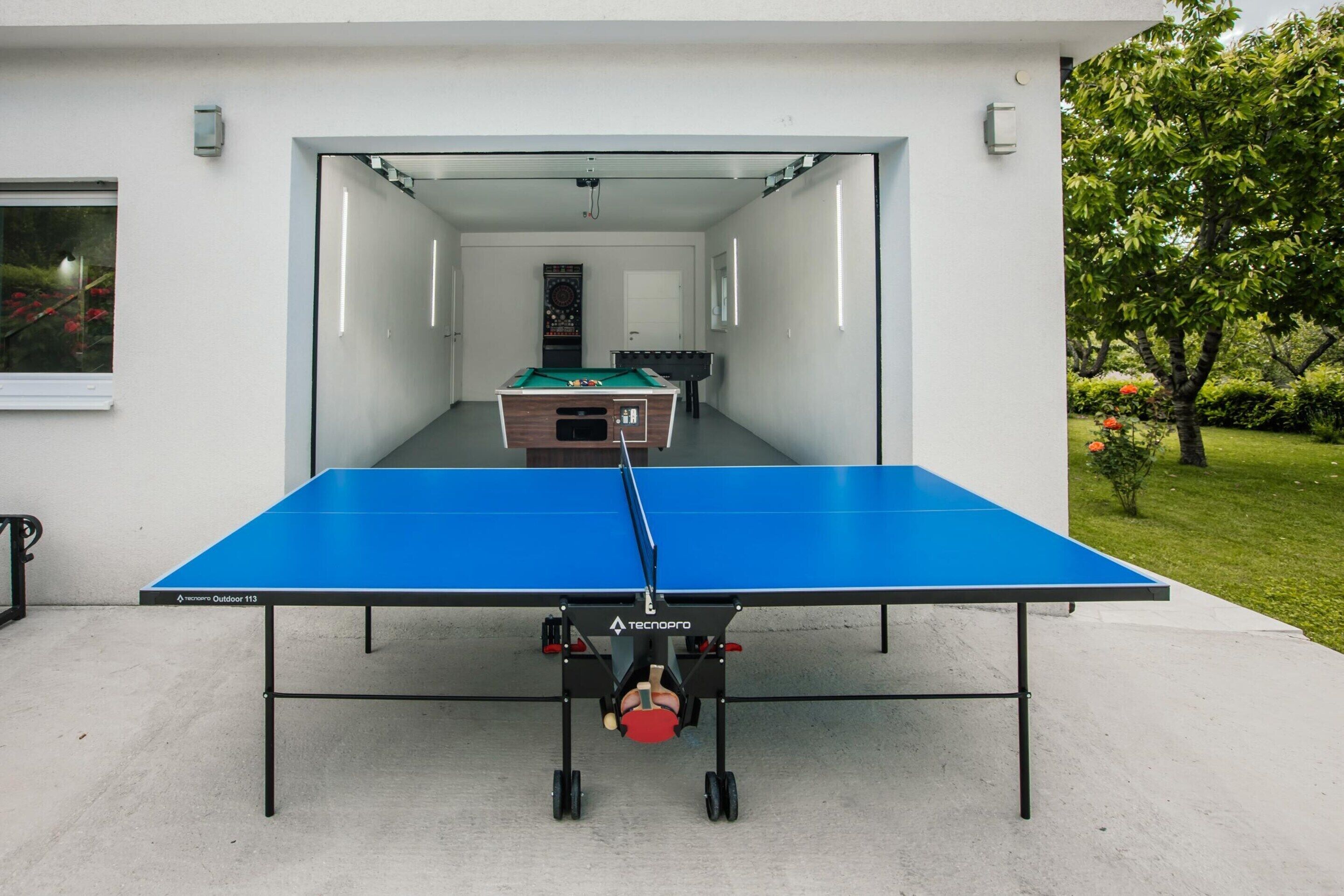 Uma mesa de pingue-pongue azul com uma rede em frente a uma parede branca, uma mesa de bilhar ao fundo e um jardim visível através de uma porta de garagem aberta.