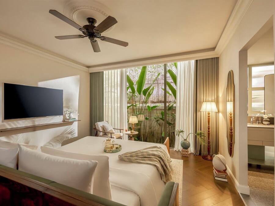 Quarto de hotel com cama, televisão, ventoinha no teto, janela com cortinas e banheiro visível por uma porta aberta.