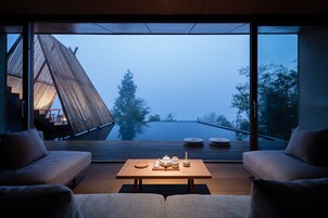 Um interior moderno com uma mesa de madeira posta com utensílios para chá, um sofá, e vista para um deck de madeira e árvores lá fora.