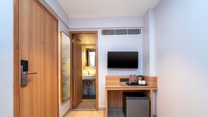 Quarto de hotel pequeno com porta de madeira, pequena área de cozinha, televisão de tela plana na parede e lixeira compacta.