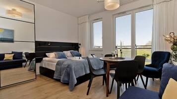 Quarto de hotel moderno com uma cama grande, uma mesa de jantar e um sofá. Janelas amplas oferecem luz natural e vista para o exterior.