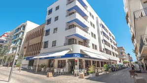 Um edifício de vários andares com um café no andar térreo, mesas ao ar livre e um céu azul claro.