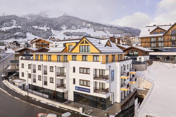 Um hotel de vários andares com varandas, um letreiro com a inscrição 'Alps Kitchen' e vista para montanhas cobertas de neve.