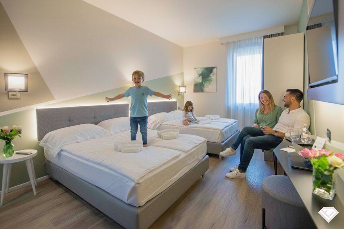 Quarto de hotel com uma cama grande, uma cama menor, uma mesa e uma cadeira. Há uma janela com cortinas, um abajur na parede e um vaso com flores na mesa.