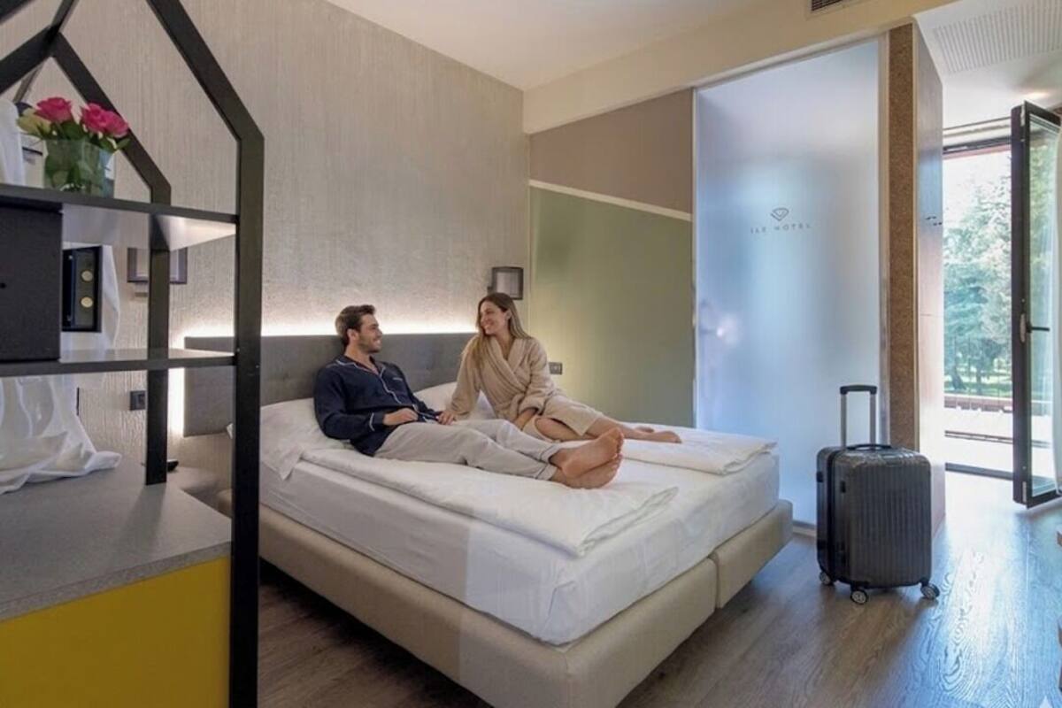 Quarto de hotel moderno com uma cama, uma mala e uma pequena mesa com um vaso de flores.