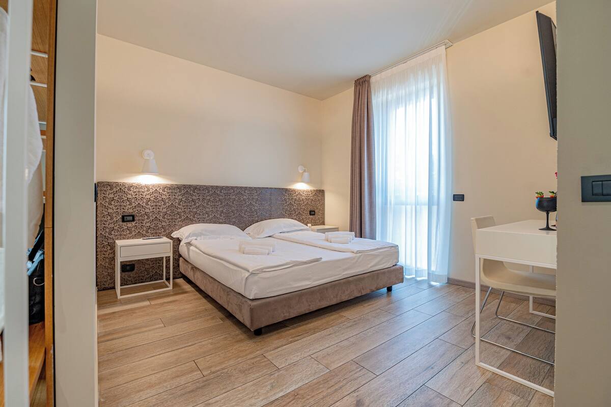 Um quarto com cama, mesas de cabeceira, escrivaninha e janela com cortinas.