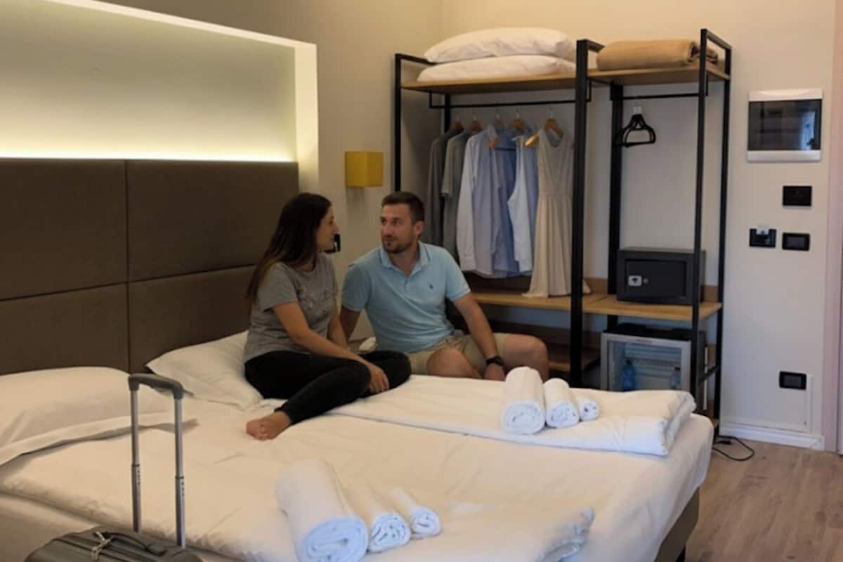 Quarto de hotel com beliche, mesa de cabeceira com abajur, mala e cama com toalhas dobradas.