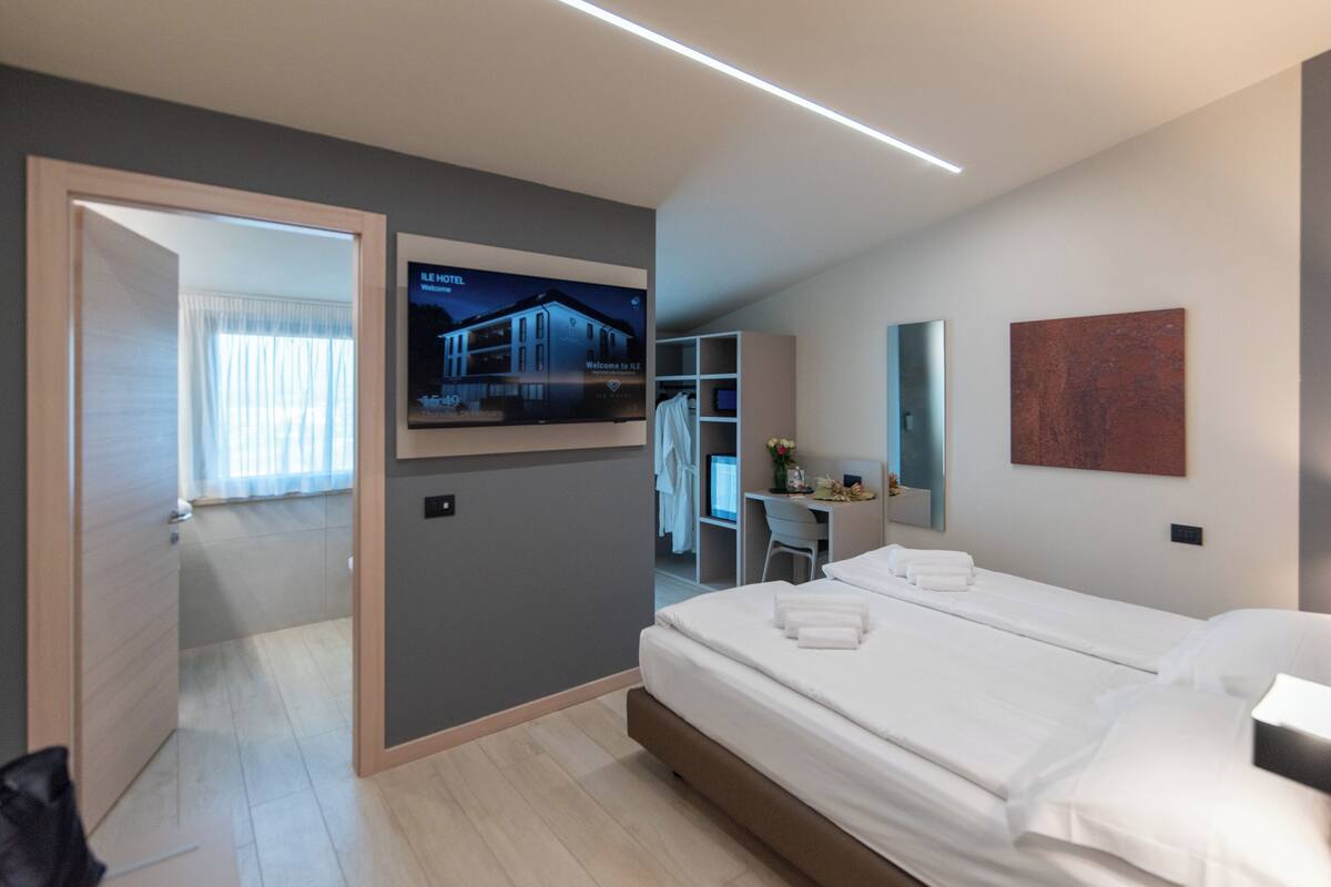 Quarto de hotel moderno com cama, escrivaninha e televisão mostrando uma imagem do hotel.