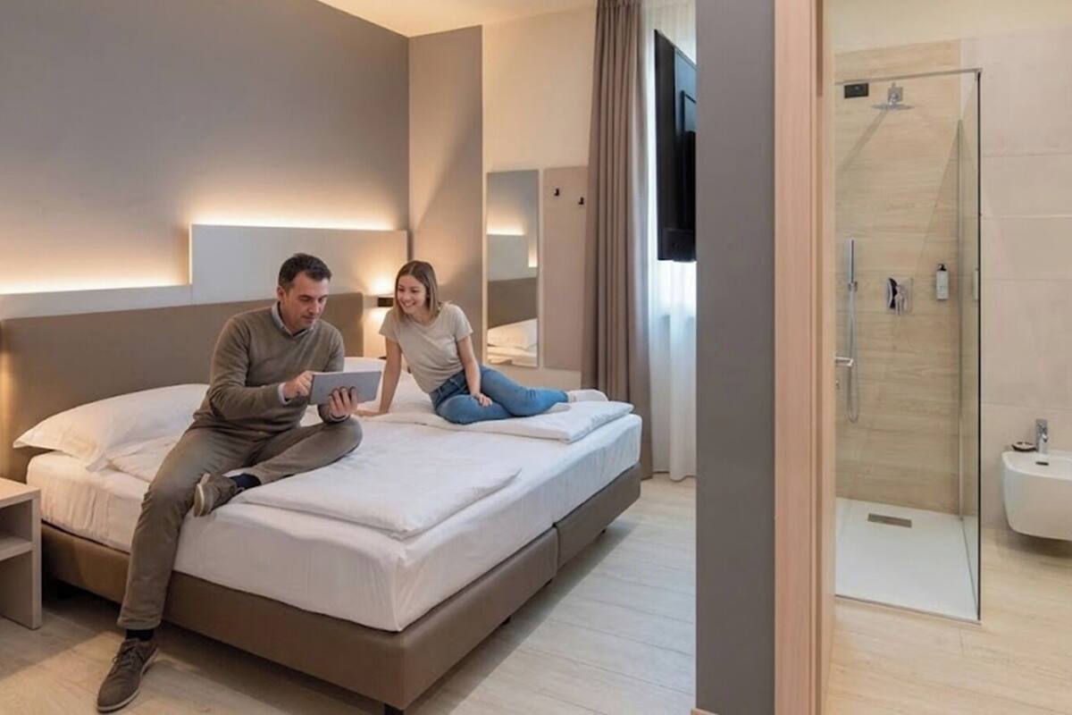 Quarto de hotel moderno com uma cama grande, mesas de cabeceira, televisão e área de chuveiro.