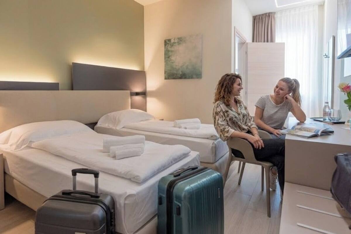 Quarto de hotel com duas camas, uma escrivaninha e uma cadeira. Há duas malas no chão.