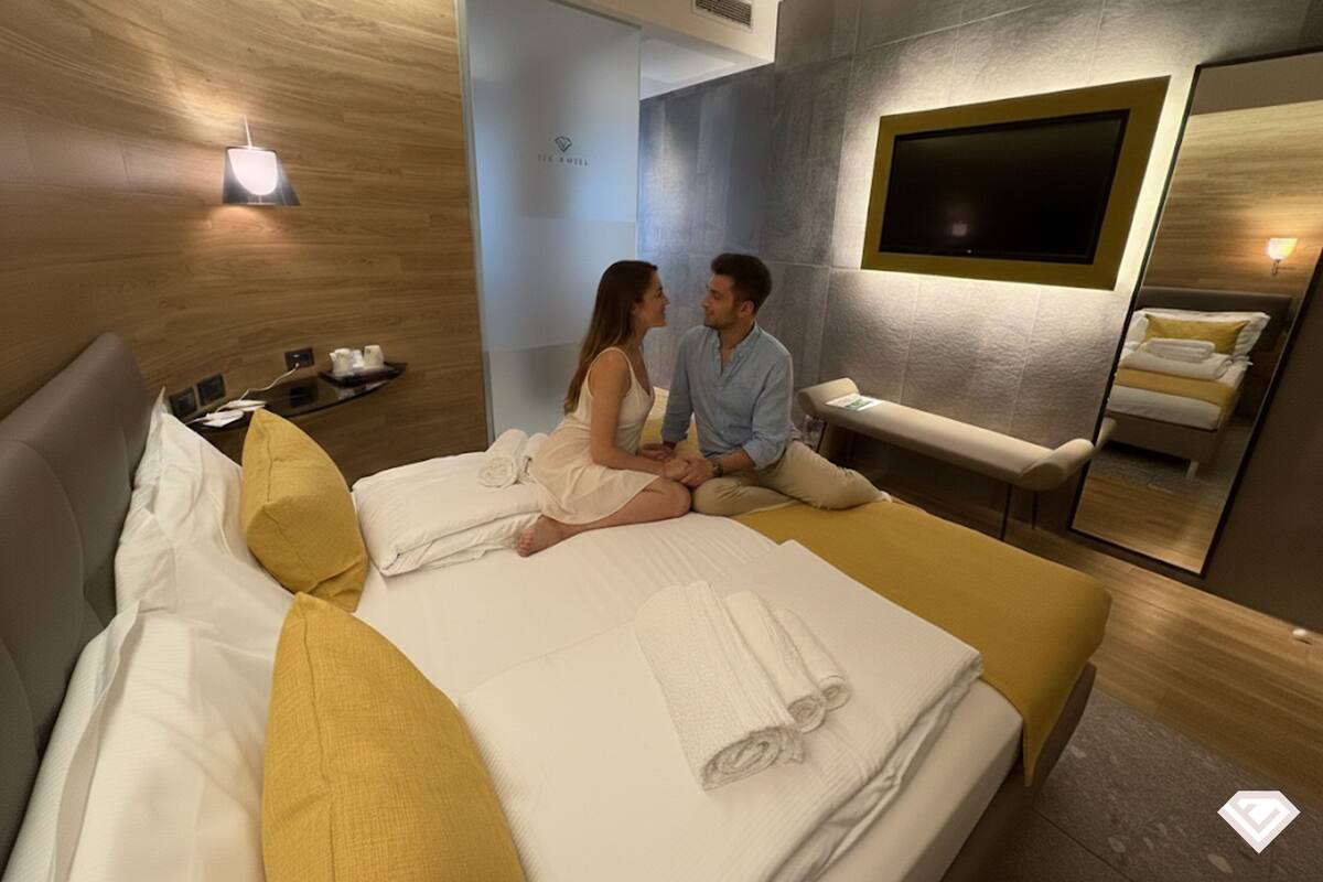 Quarto de hotel moderno com uma cama grande, mesa de cabeceira, espelho e televisão fixada na parede.