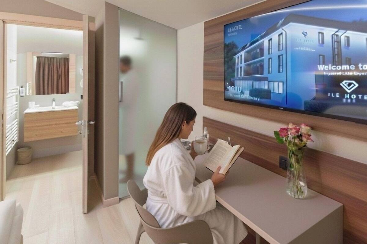 Quarto de hotel moderno com uma televisão de tela plana grande exibindo uma mensagem de boas-vindas, uma escrivaninha de madeira, uma cadeira e um vaso com flores.
