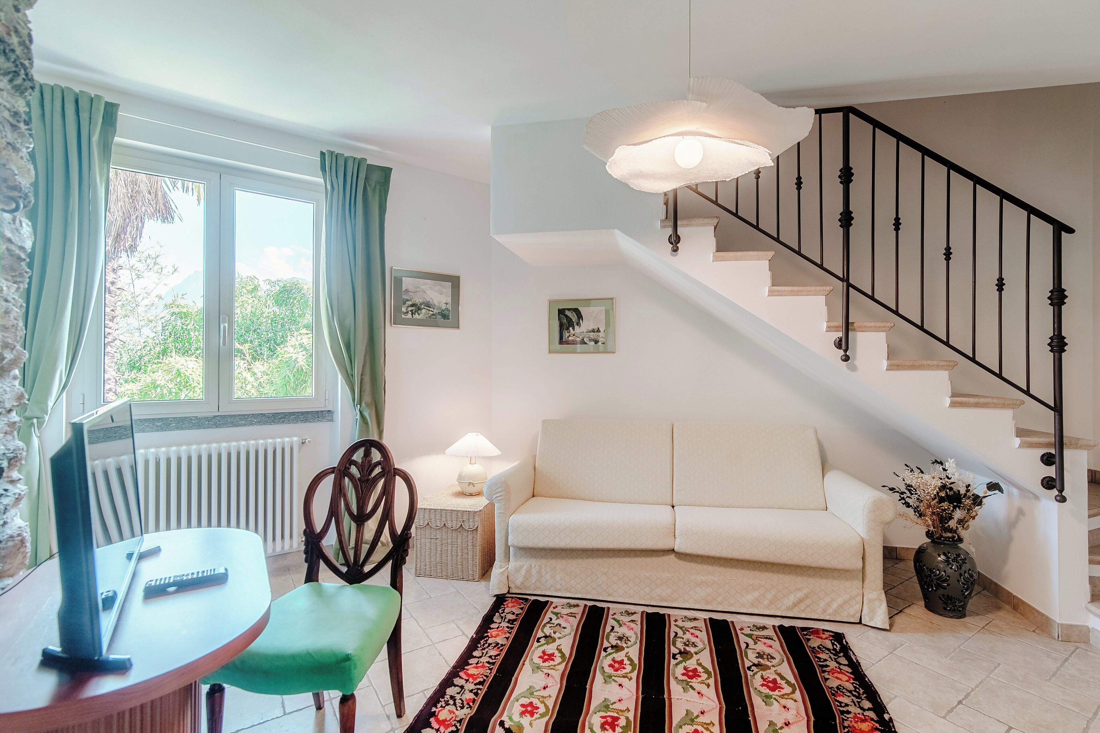 Uma sala de estar com sofá, uma mesa de jantar com cadeiras, escada e janela com cortinas.