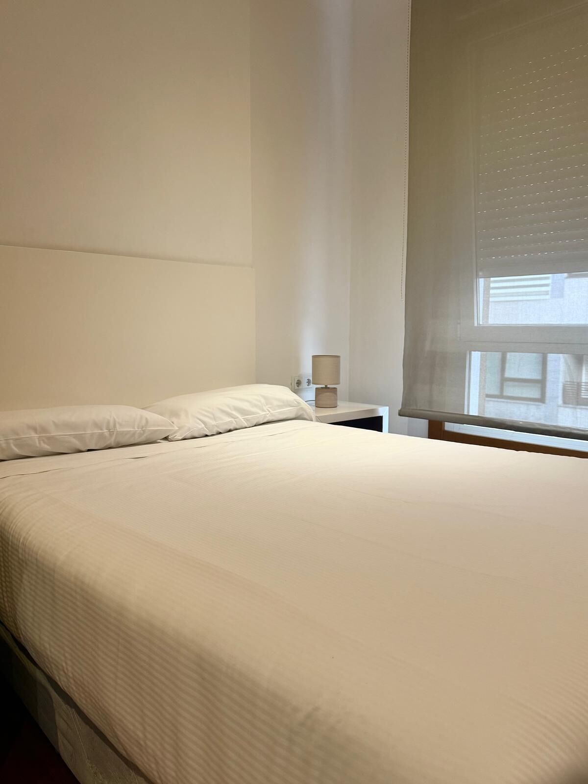 Cama com roupa de cama branca em quarto com janela e mesa de cabeceira com abajur.