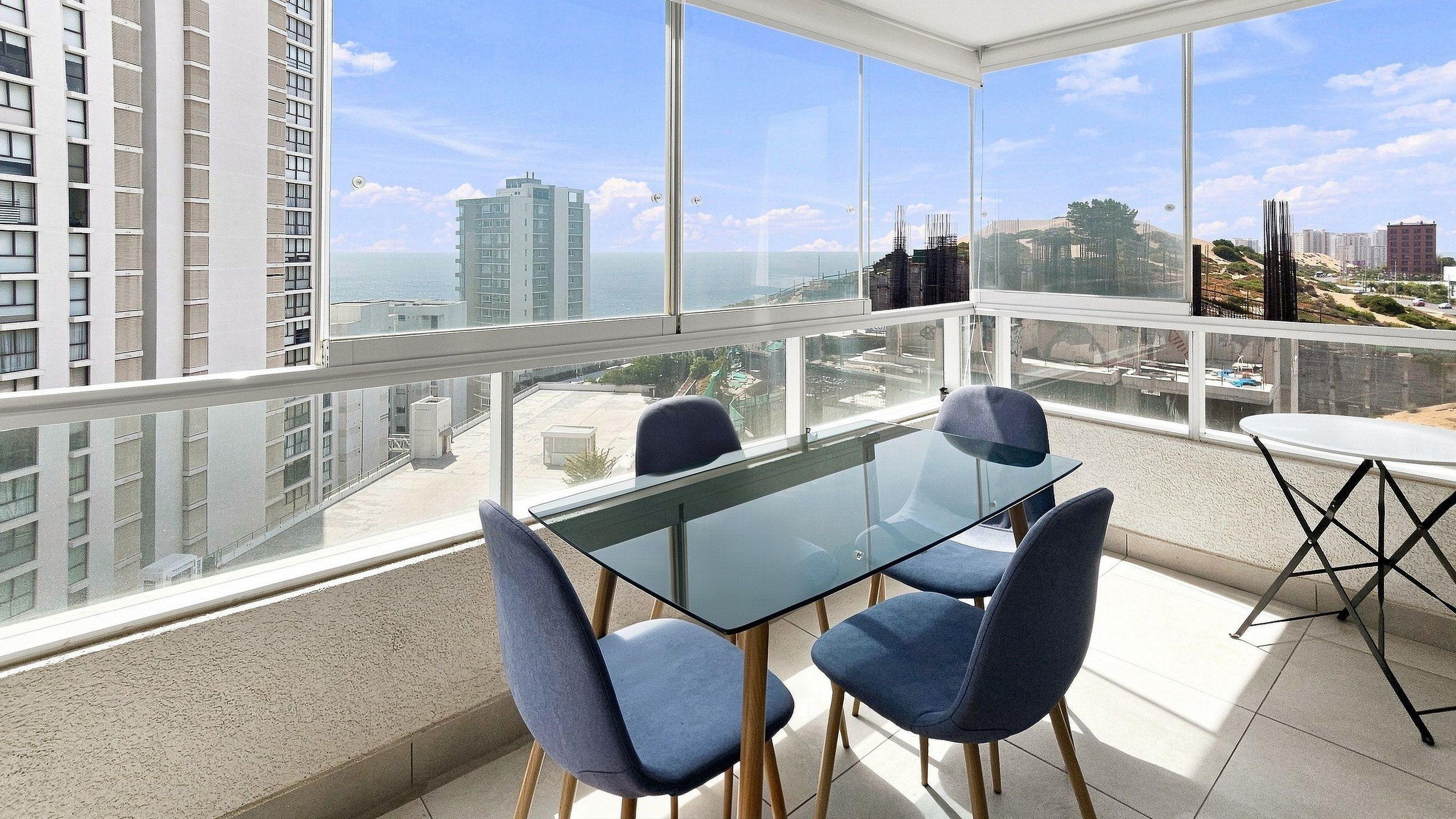 Quarto moderno com mesa e cadeiras de vidro, grandes janelas com vista para a cidade e uma varanda.