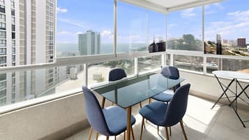 Quarto moderno com mesa e cadeiras de vidro, grandes janelas com vista para a cidade e uma varanda.