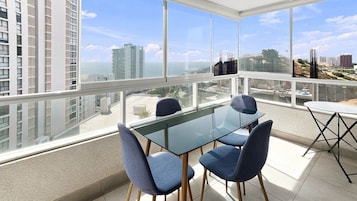 Quarto moderno com mesa e cadeiras de vidro, grandes janelas com vista para a cidade e uma varanda.