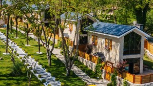 Um hotel moderno com fachada de pedra e madeira, rodeado por vegetação exuberante e um caminho de acesso pavimentado.