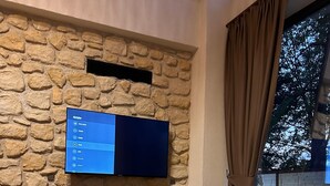 Paredede pedra com uma TV de tela plana montada sobre uma lareira. Uma mesa de madeira com um controle remoto está em primeiro plano.
