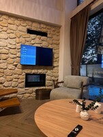 Paredede pedra com uma TV de tela plana montada sobre uma lareira. Uma mesa de madeira com um controle remoto está em primeiro plano.