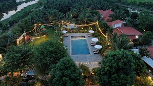 Um resort com piscina rodeada por palmeiras e luzes de corda, situado numa paisagem exuberante com um rio ao fundo.