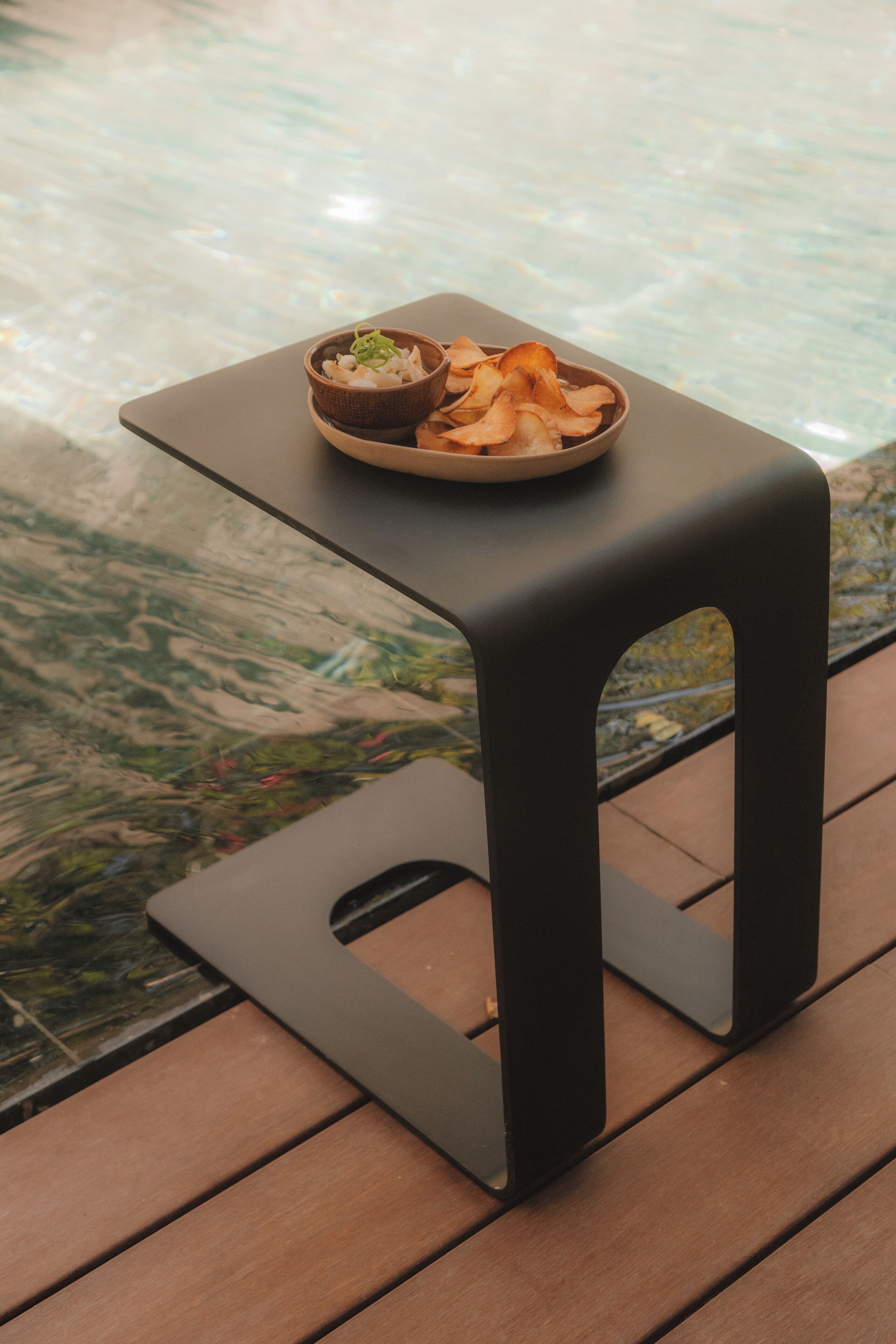 Um banco preto com uma tigela de comida sobre ele, colocado sobre um deck de madeira perto de uma superfície refletora.