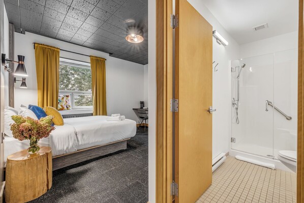 Quarto de hotel com cama, secretária, cadeira, janela com cortinas e zona de chuveiro.