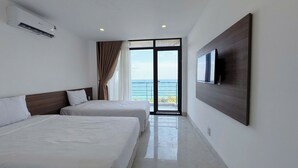 Quarto de hotel com duas camas, uma televisão grande na parede e vista para o oceano por uma porta de correr.