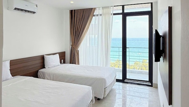 Quarto de hotel com duas camas, televisão, ar condicionado e uma grande janela vista para o mar.