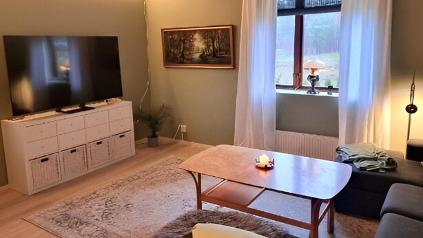 Uma sala de estar com televisão, um quadro, uma janela com cortinas, uma mesa e um sofá.