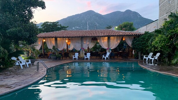 Piscina com cadeiras brancas e área de estar coberta, cercada por vegetação e montanhas ao fundo.