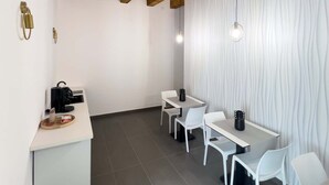 Quarto moderno e minimalista com vigas de madeira no teto, paredes brancas e um painel de parede branco texturizado. Há mesas e cadeiras brancas dispostas para refeições ou trabalho.