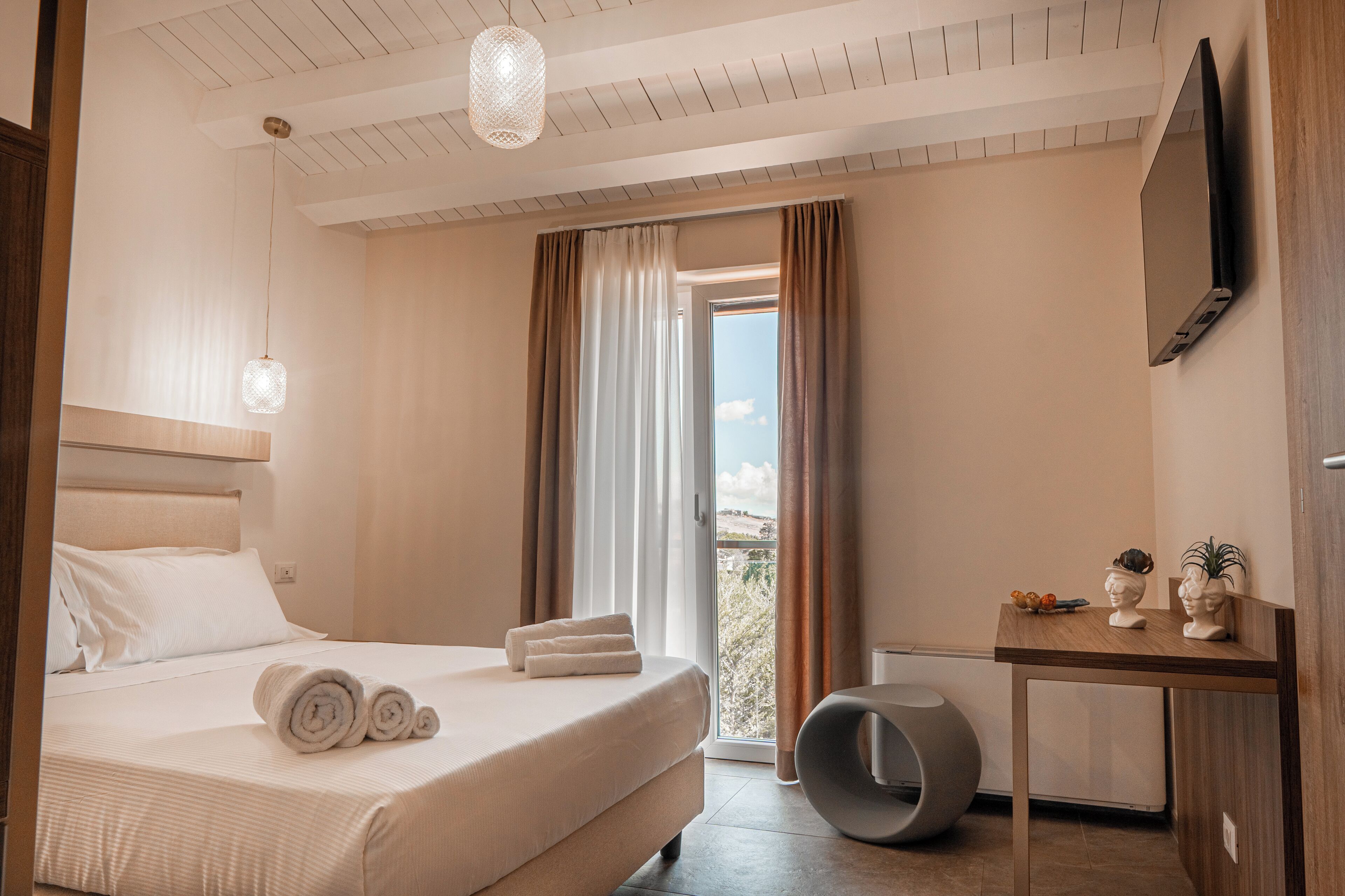 Quarto de hotel com cama, um espaço de estar circular, uma mesa com objetos decorativos e uma TV fixada na parede.