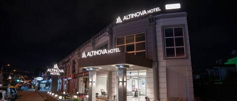 Exterior do Hotel Altinova à noite, com entrada bem iluminada, degraus que levam à porta, área de recepção visível e estacionamento com carros.