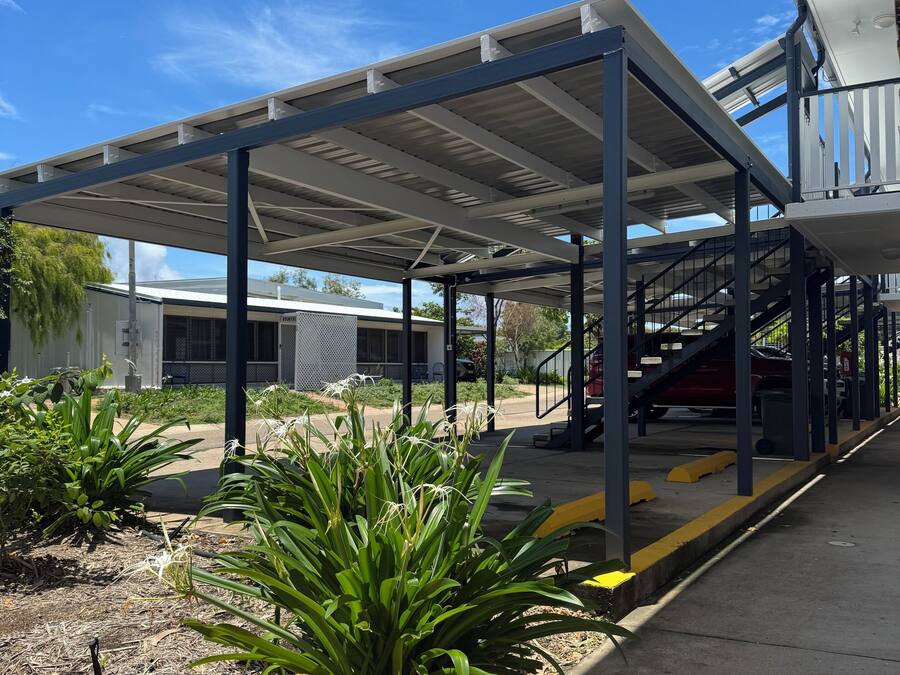 Uma área externa coberta com telhado de metal, piso de concreto e plantas em vasos.