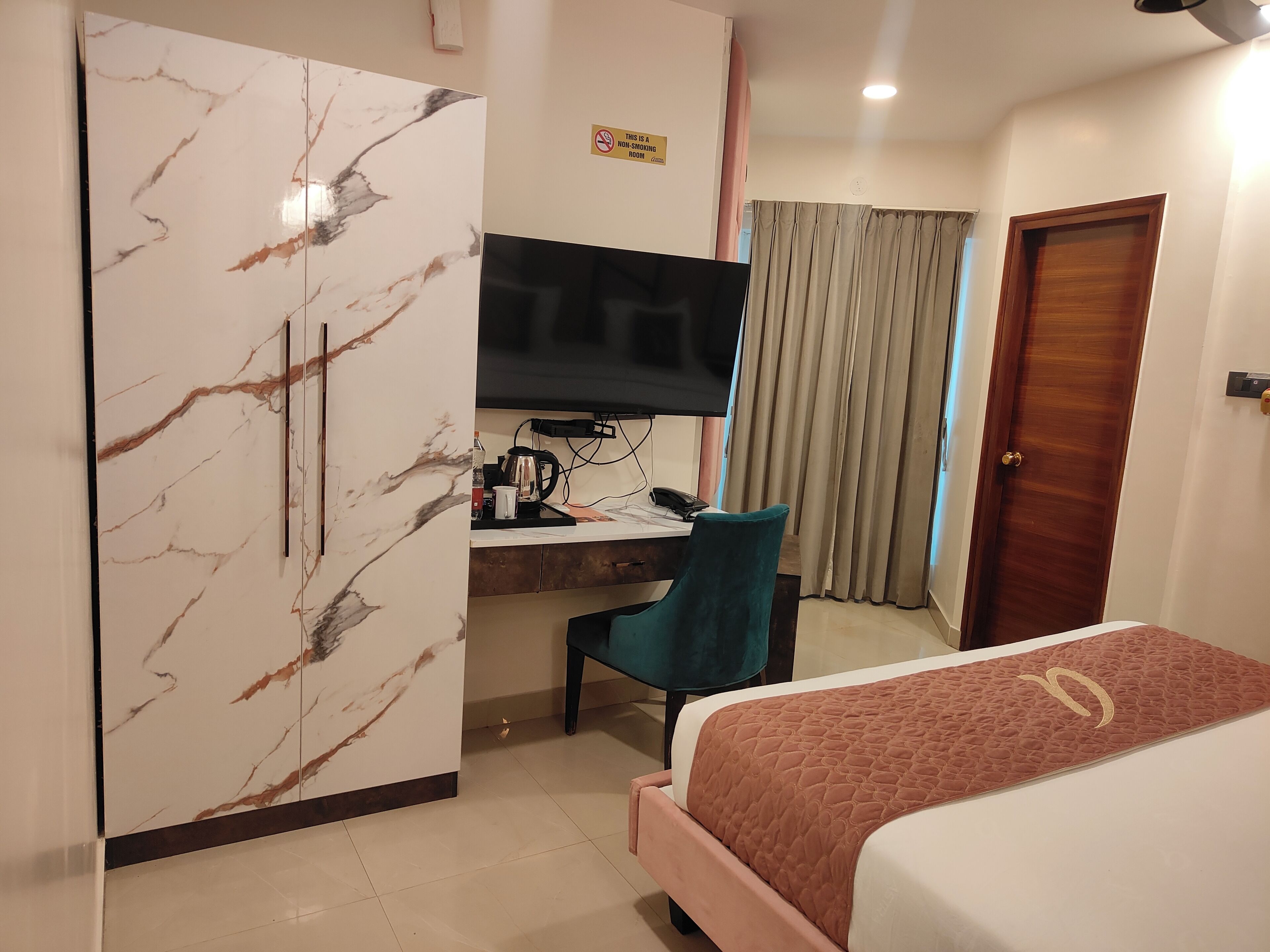Quarto de hotel com parede de mármore, cama com colcha marrom, escrivaninha com televisão, cadeira e porta de madeira.