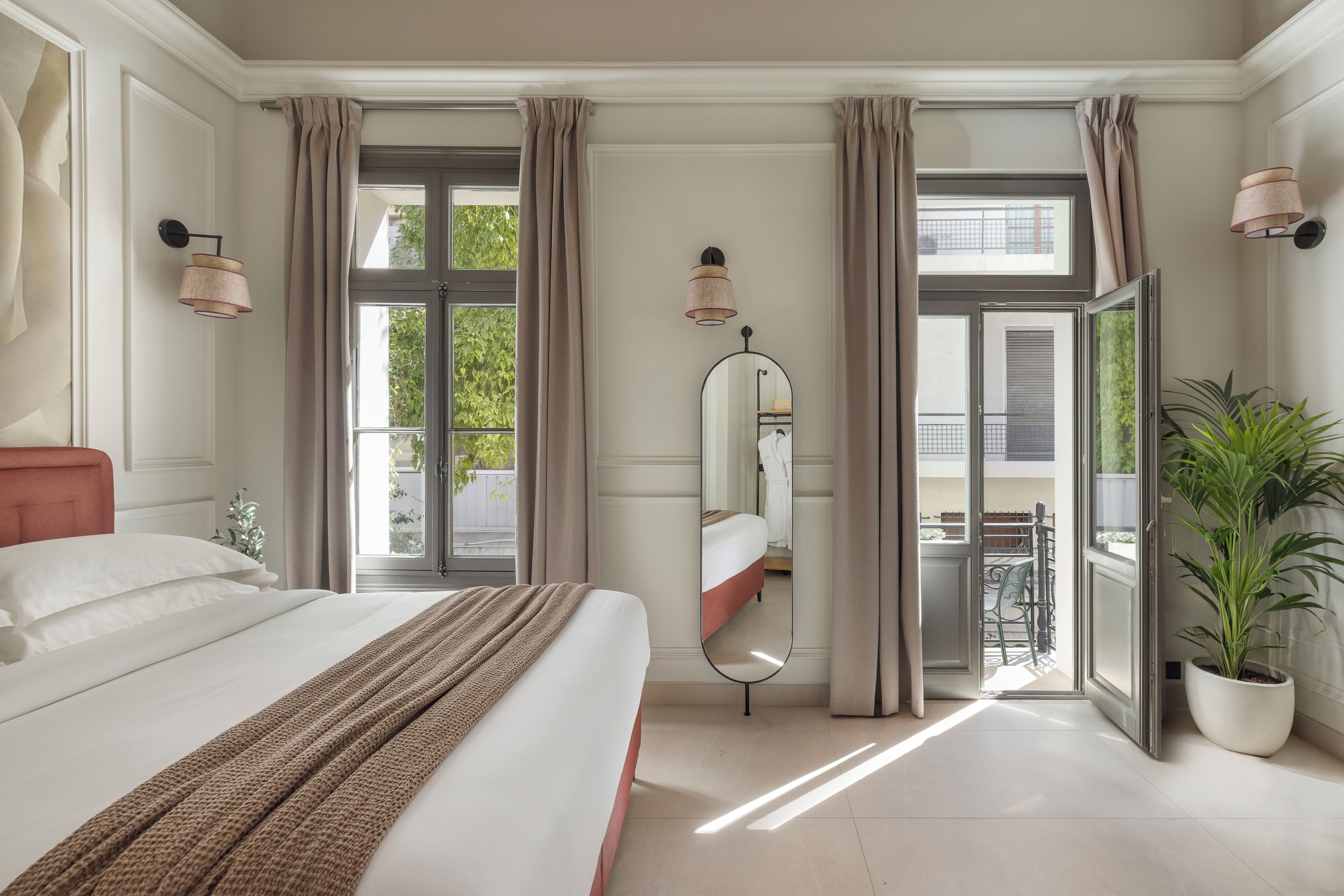 Quarto de hotel com cama, um espelho grande, uma planta em vaso e vista para o exterior.