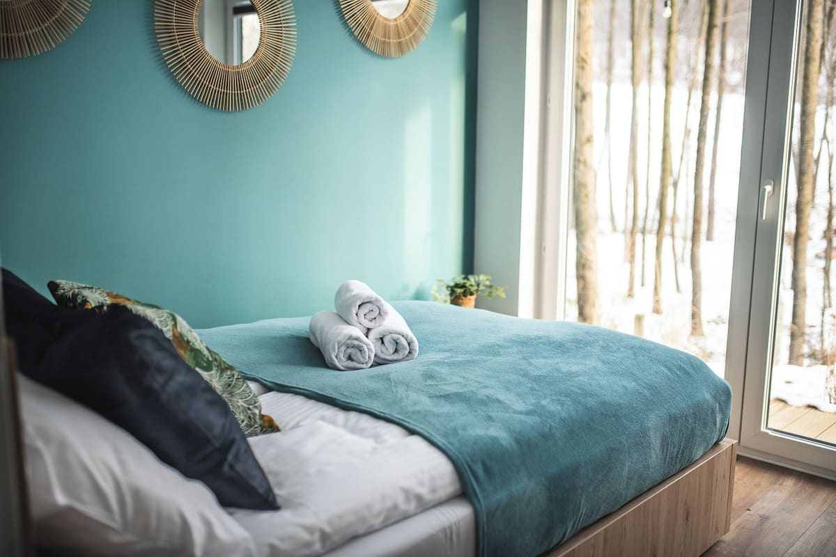 Uma cama com colcha verde-água, almofadas brancas e toalhas dobradas. Há dois espelhos na parede e um pequeno vaso com planta no parapeito da janela.