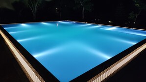 Piscina retangular com entorno escuro e bordas iluminadas, rodeada por árvores.