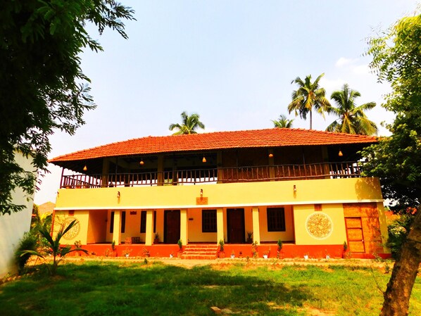 Um edifício de dois andares com telhado de telhas vermelhas, várias varandas e um jardim com palmeiras.