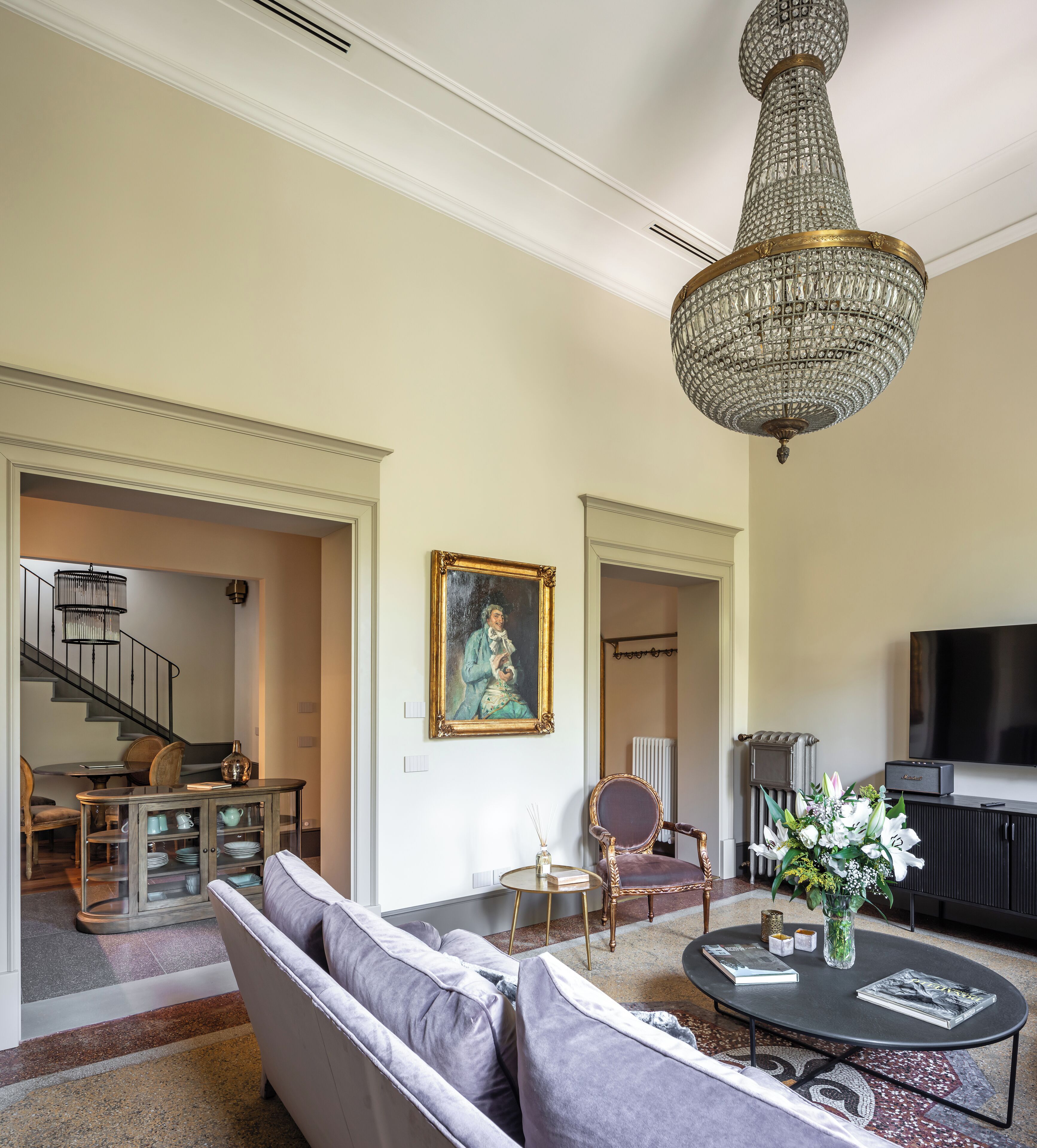 Uma sala de estar com um candelabro, um sofá, uma mesa de centro, um quadro, uma poltrona, uma lareira e uma televisão.