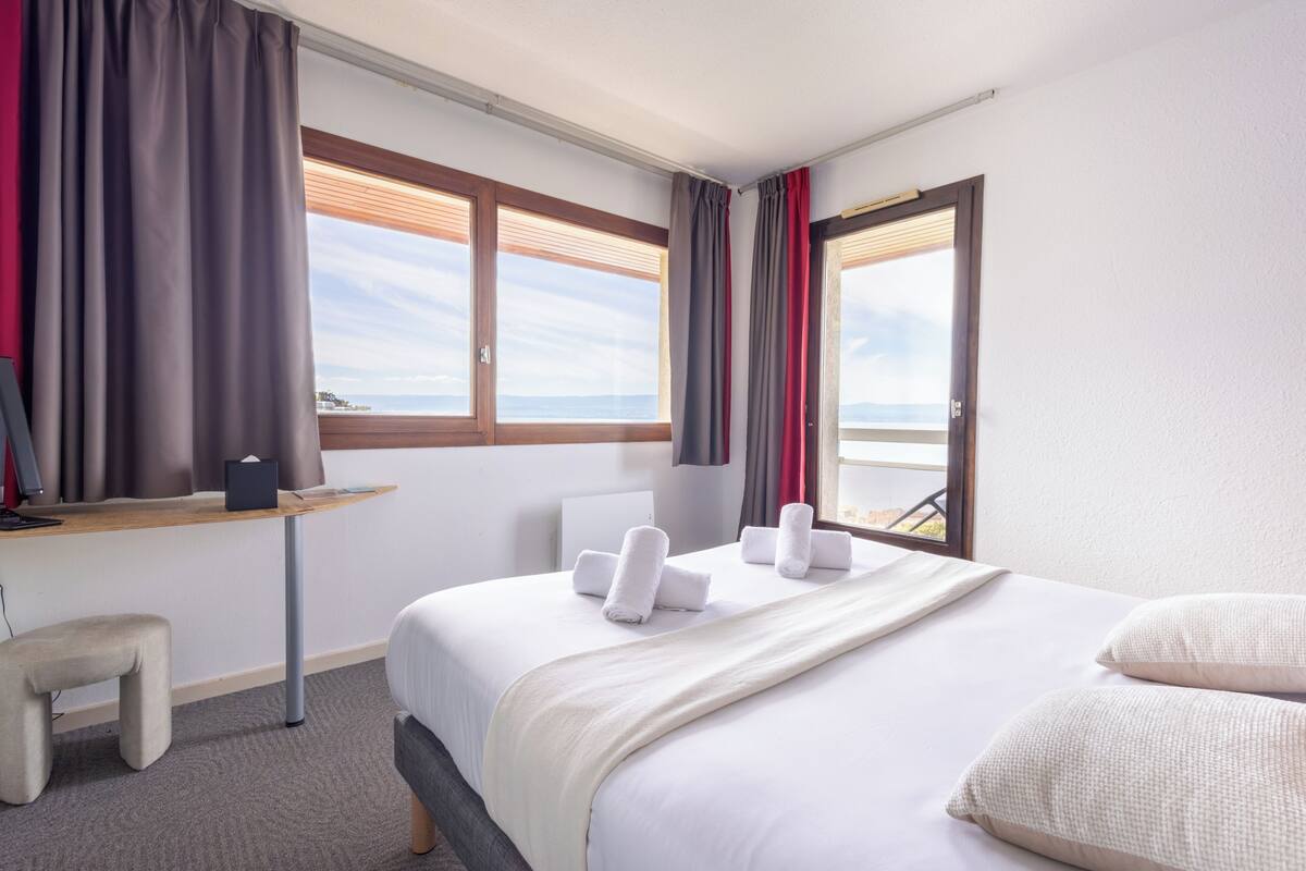 Quarto de hotel com cama, escrivaninha, cadeira e janela com cortinas.