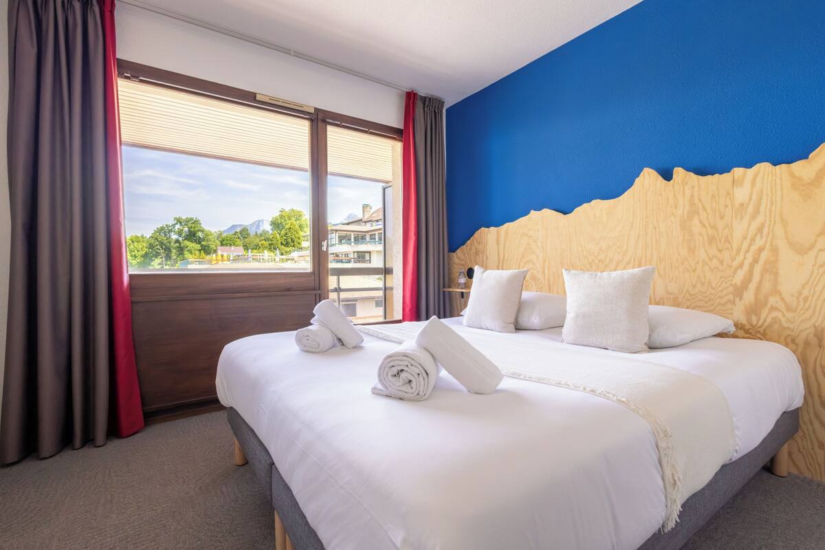 Quarto de hotel com uma cama grande, cabeceira de madeira e vista para edifícios e árvores pela janela.