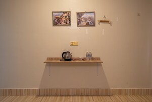 Um quarto com piso de madeira, uma estante com uma chaleira, duas garrafas e duas fotos emolduradas na parede.