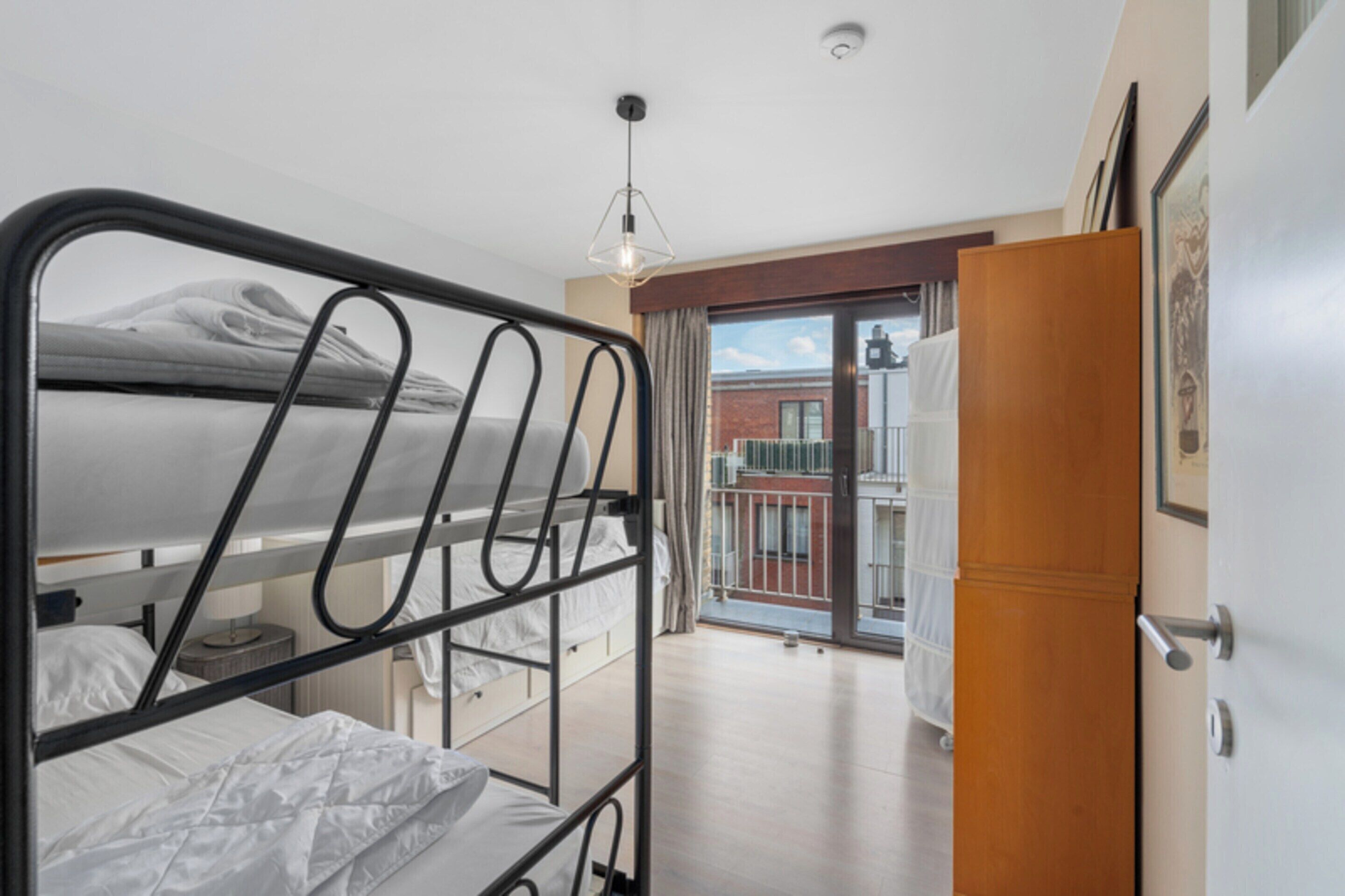 Quarto com beliches, porta de madeira e vista para um edifício através de uma porta de vidro.
